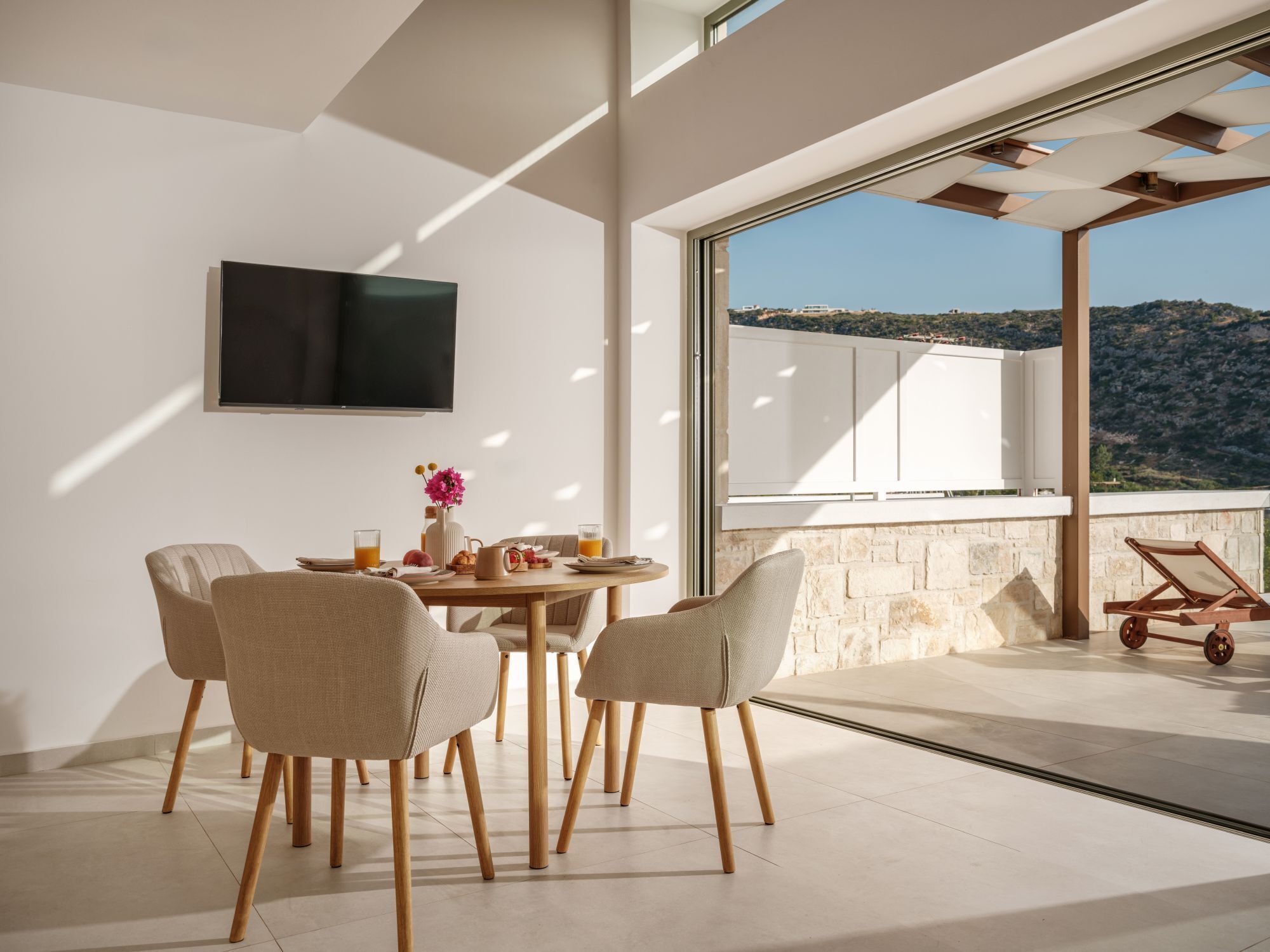 Uma sala de jantar moderna com uma mesa posta para quatro pessoas, uma TV de tela plana na parede e uma janela grande com vista para o campo.