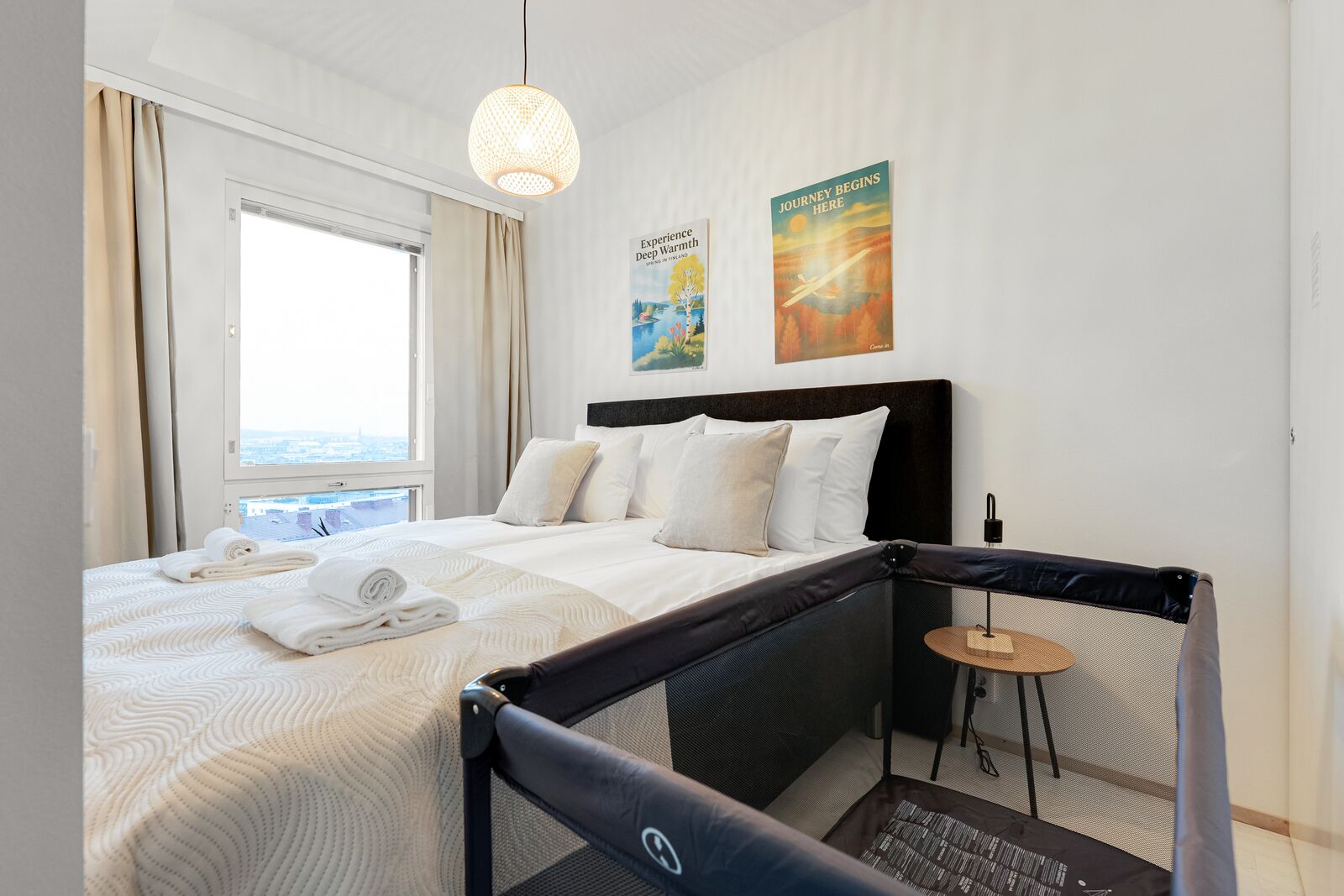 Quarto de hotel com uma cama, janela, luminária pendente e dois cartazes na parede.