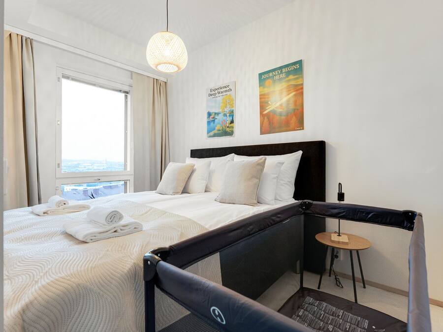 Quarto de hotel com uma cama, janela, luminária pendente e dois cartazes na parede.