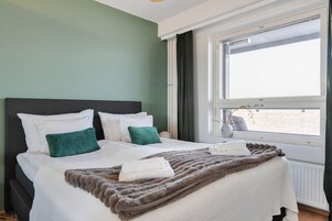 Uma cama bem arrumada com travesseiros brancos e verdes, um cobertor marrom fofinho e uma janela com vista para uma paisagem nevada.