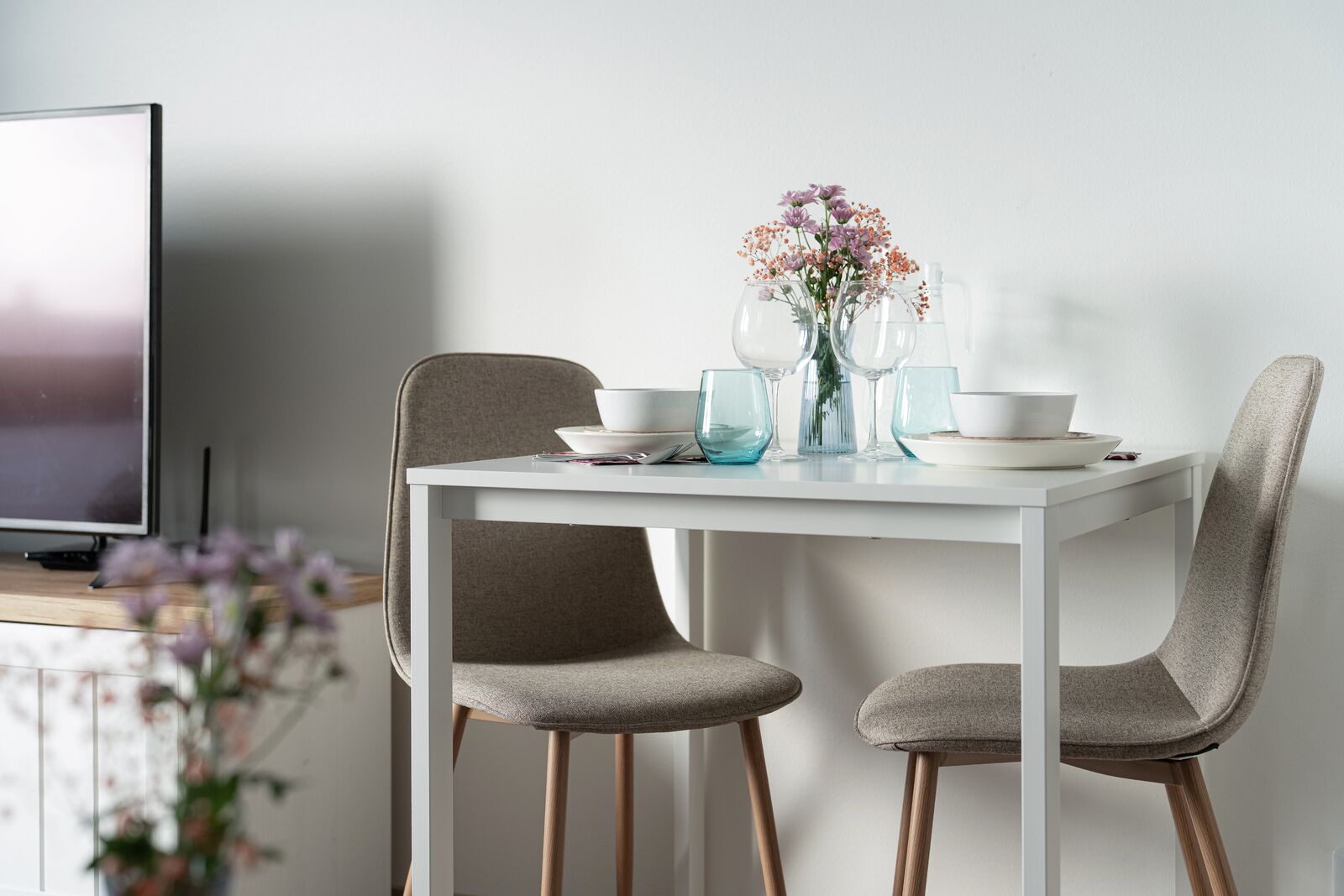 Uma área de refeições com mesa branca, duas cadeiras cinzas, um vaso com flores e uma televisão sobre um suporte.