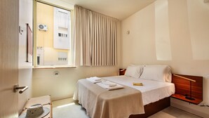Quarto de hotel com cama, mesa de cabeceira com chapéu, janela com cortinas e ar condicionado fixado na parede.