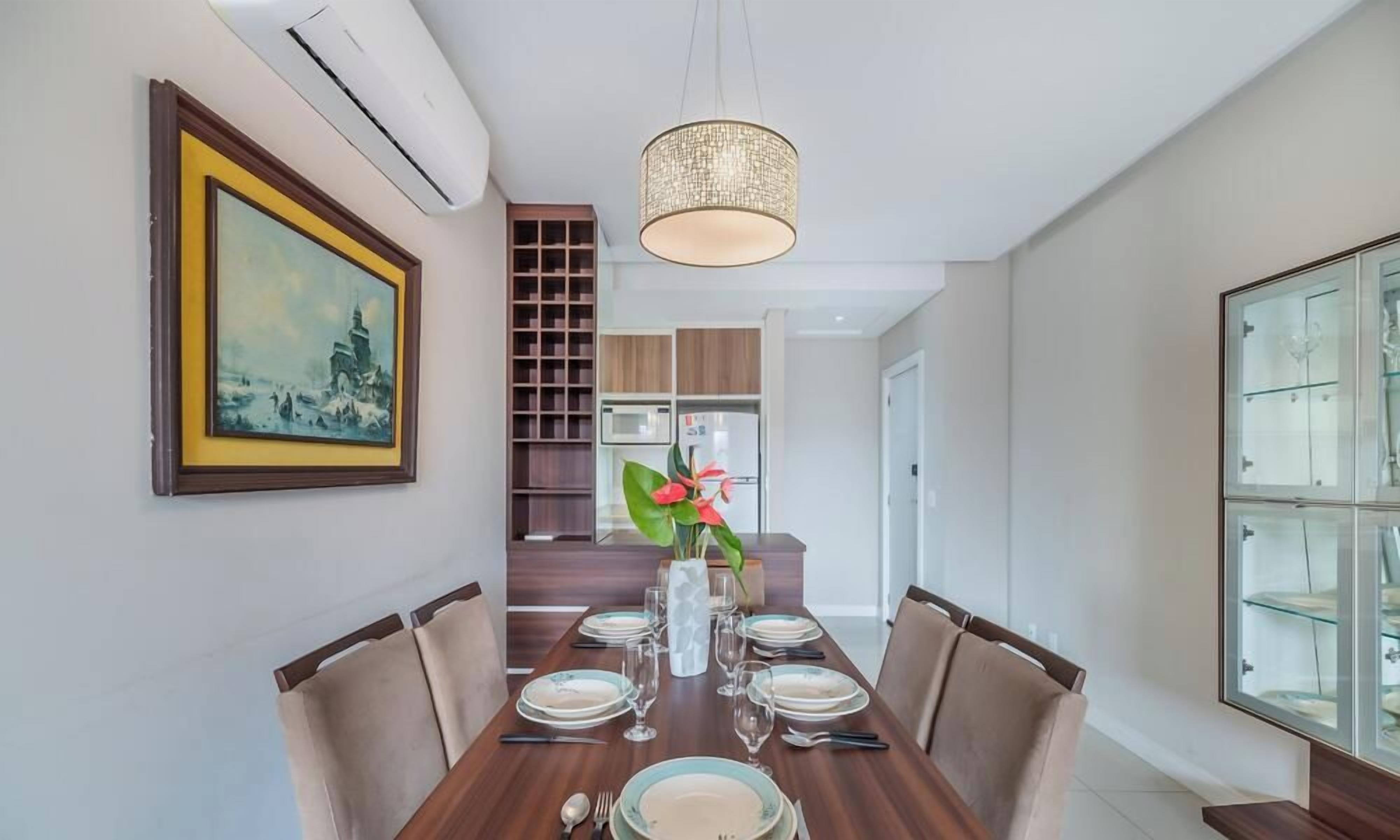 Uma sala de jantar com mesa de madeira posta para quatro pessoas, um vaso com flores e um quadro grande na parede.
