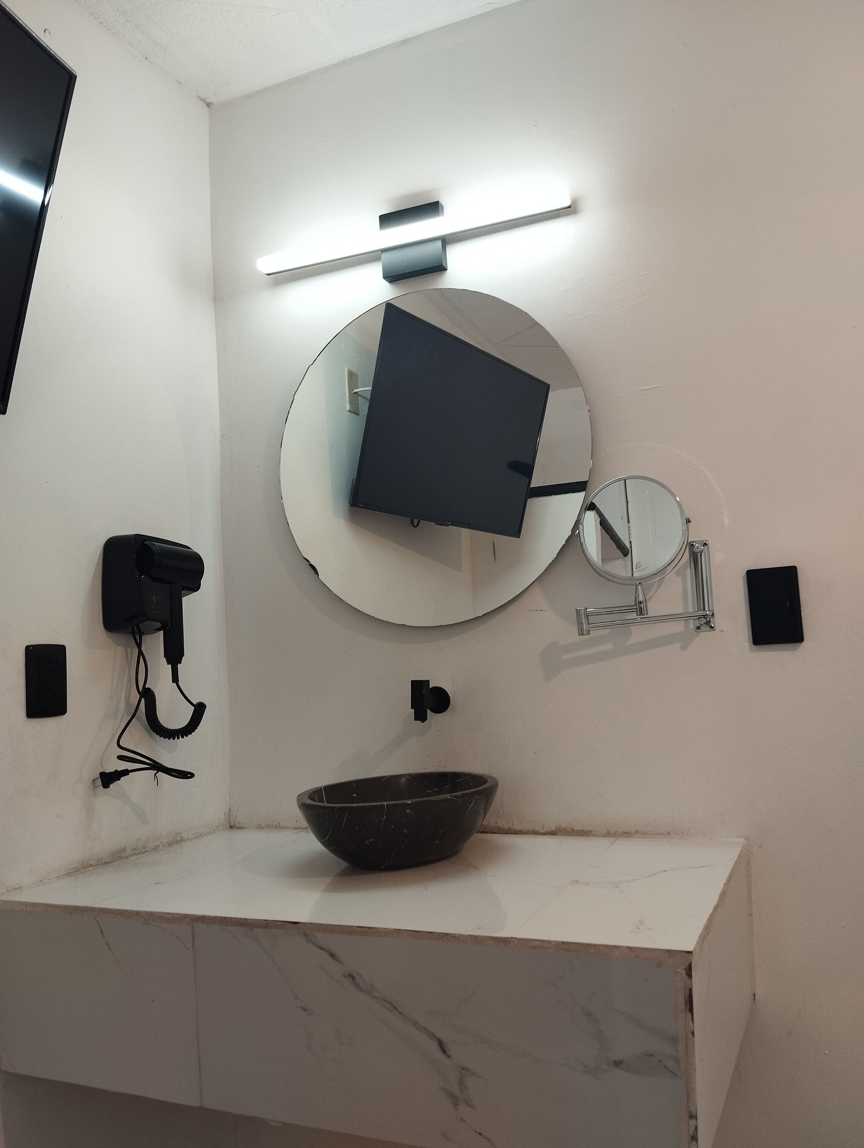 Uma casa de banho com lavatório de mármore branco, espelho redondo, telefone preto e luminária fixada na parede.