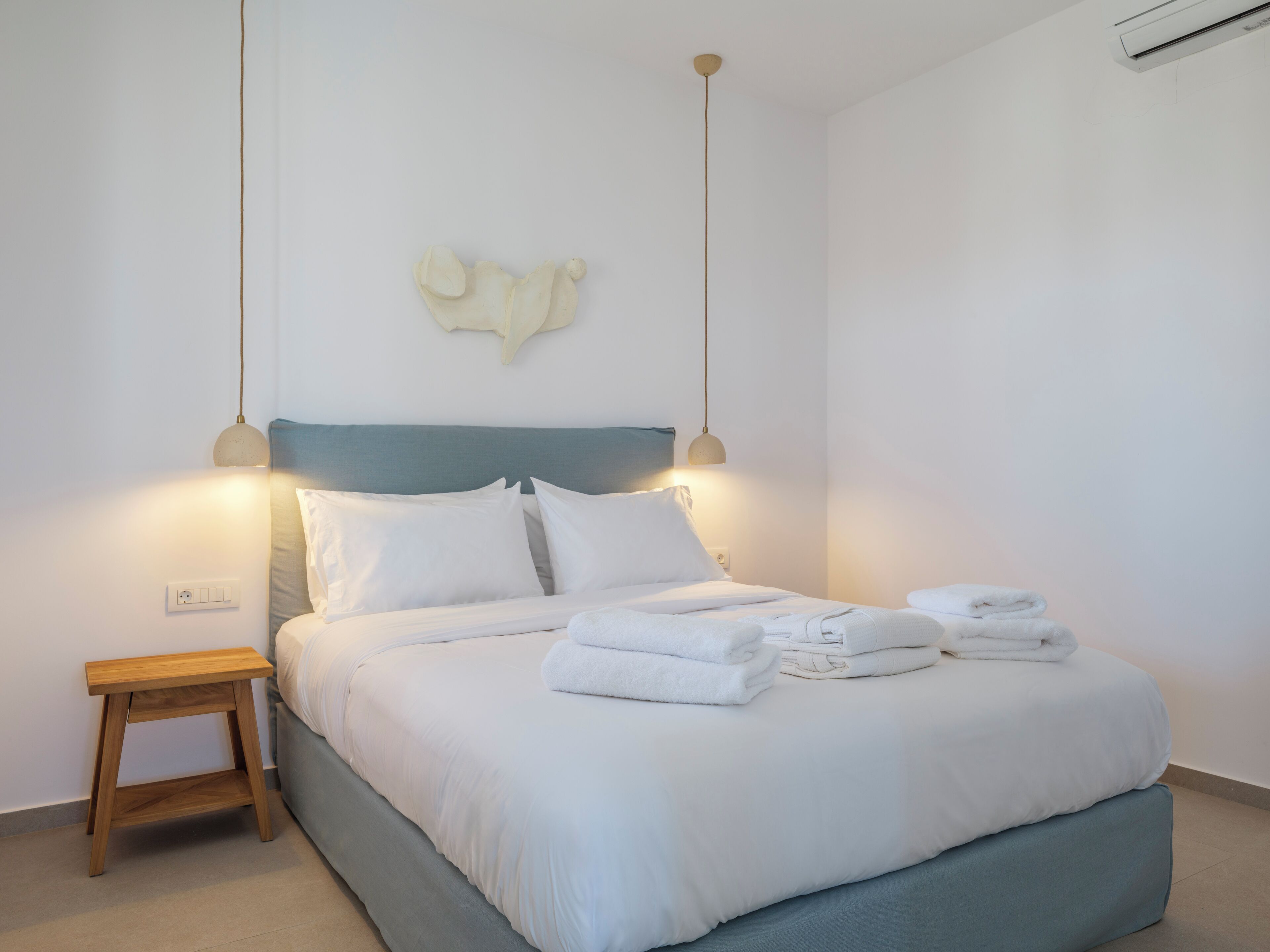 Cama bem arrumada com lençóis brancos e cabeceira azul, um pequeno criado-mudo de madeira e luminária fixada na parede.
