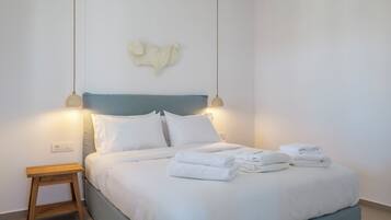 Cama bem arrumada com lençóis brancos e cabeceira azul, um pequeno criado-mudo de madeira e luminária fixada na parede.