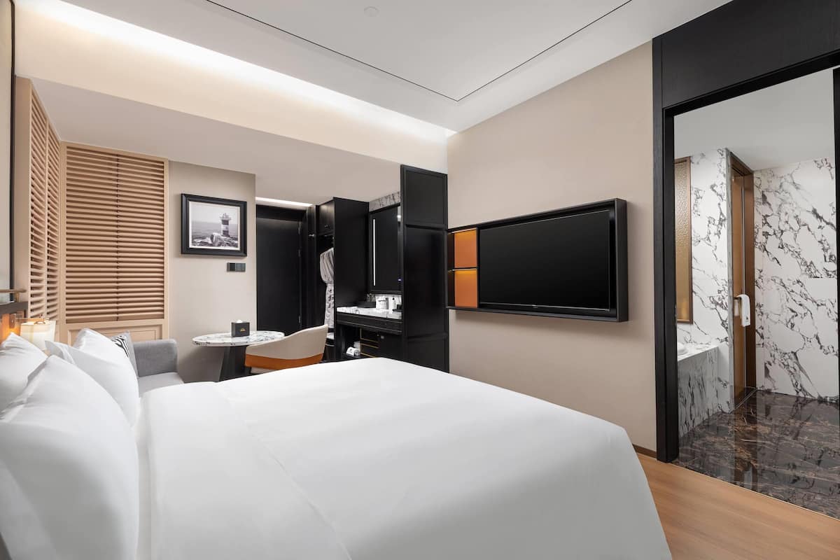 Quarto de hotel moderno com uma cama grande, uma escrivaninha, uma cadeira, uma televisão e um banheiro com azulejos de mármore.