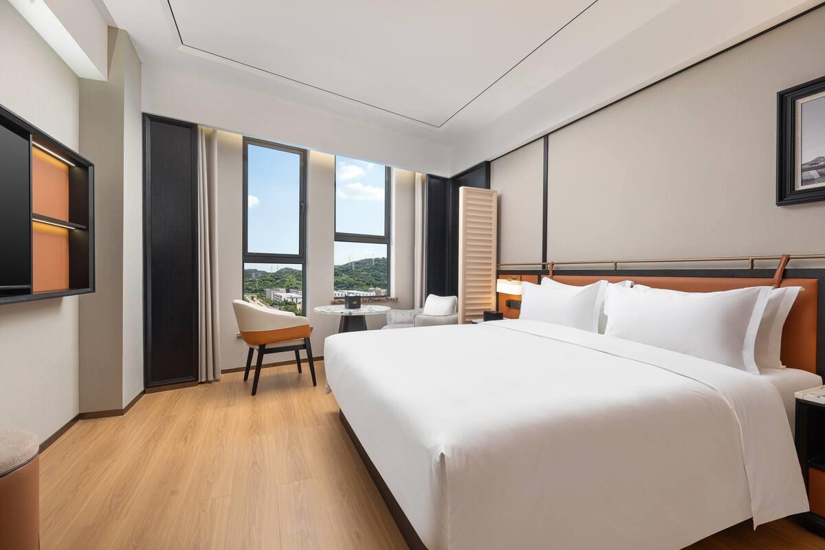 Quarto de hotel moderno com uma cama grande, uma escrivaninha e uma cadeira. Há uma janela com vista para a cidade.