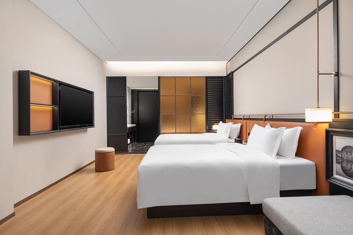 Quarto de hotel com duas camas, uma televisão de tela plana na parede, um pequeno banquinho e um quadro emoldurado.