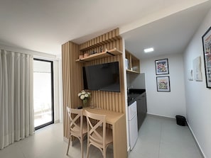 Espaço compacto com cozinha compacta, televisão de tela plana fixada em um painel de madeira, duas cadeiras, mesa pequena para refeições e porta de vidro deslizante que dá para uma área externa.