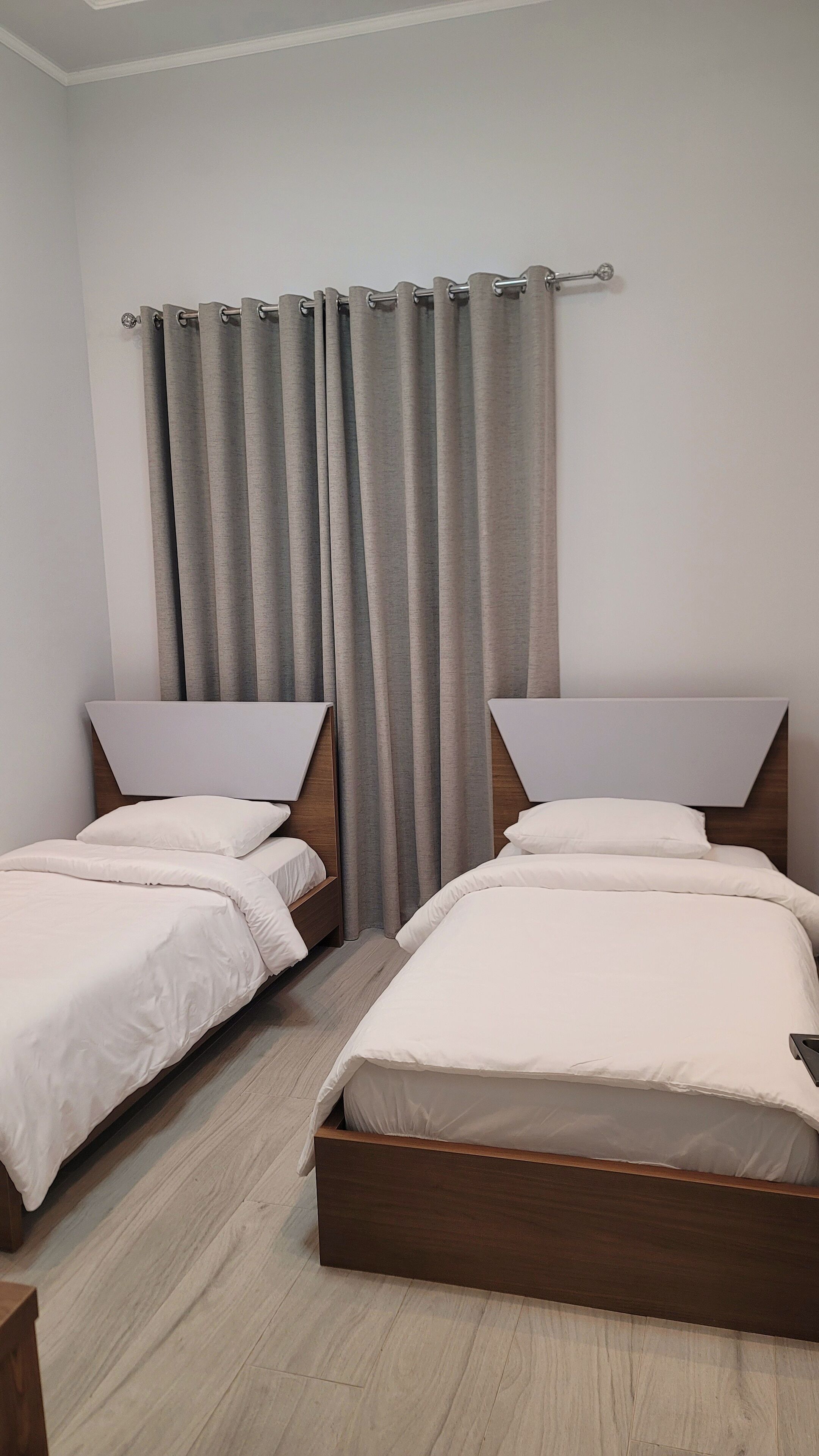 Quarto de hotel com duas camas de solteiro, cabeceira de madeira, cortina cinza e piso de madeira.