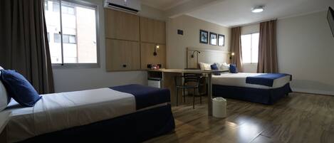 Quarto de hotel compacto com duas camas de solteiro, uma kitchenette pequena, uma escrivaninha e uma cadeira.