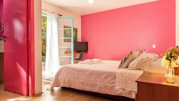 Quarto com parede rosa, cama com roupa de cama branca, mesa de cabeceira de madeira e TV sobre suporte.