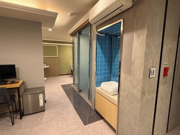 Um banheiro de hotel com chuveiro, pia e vaso sanitário. Há uma escrivaninha com um computador e uma lixeira.