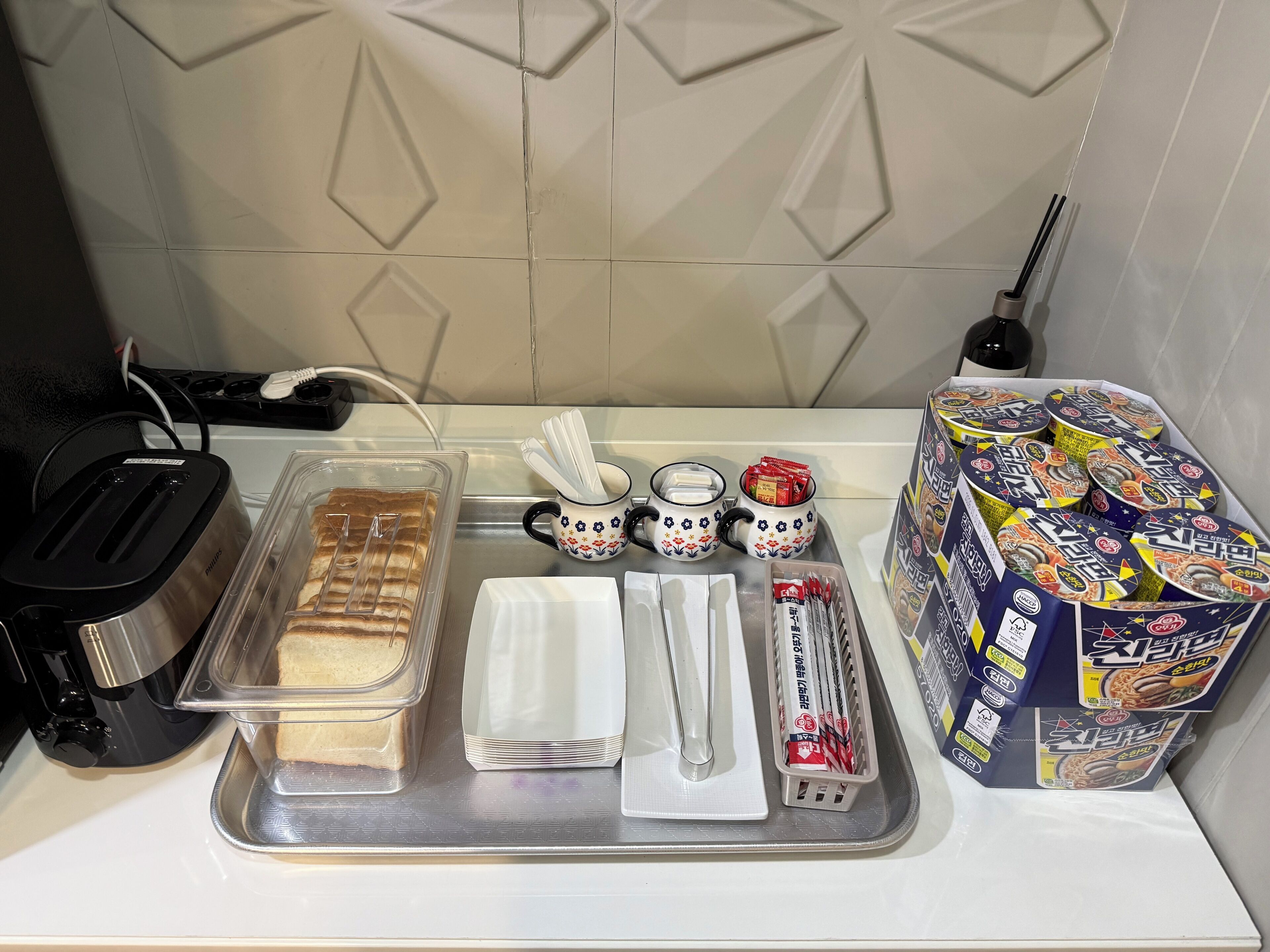 Bandeja de café da manhã com pão, utensílios, uma caneca, uma torradeira e uma caixa de macarrão instantâneo em uma bancada.