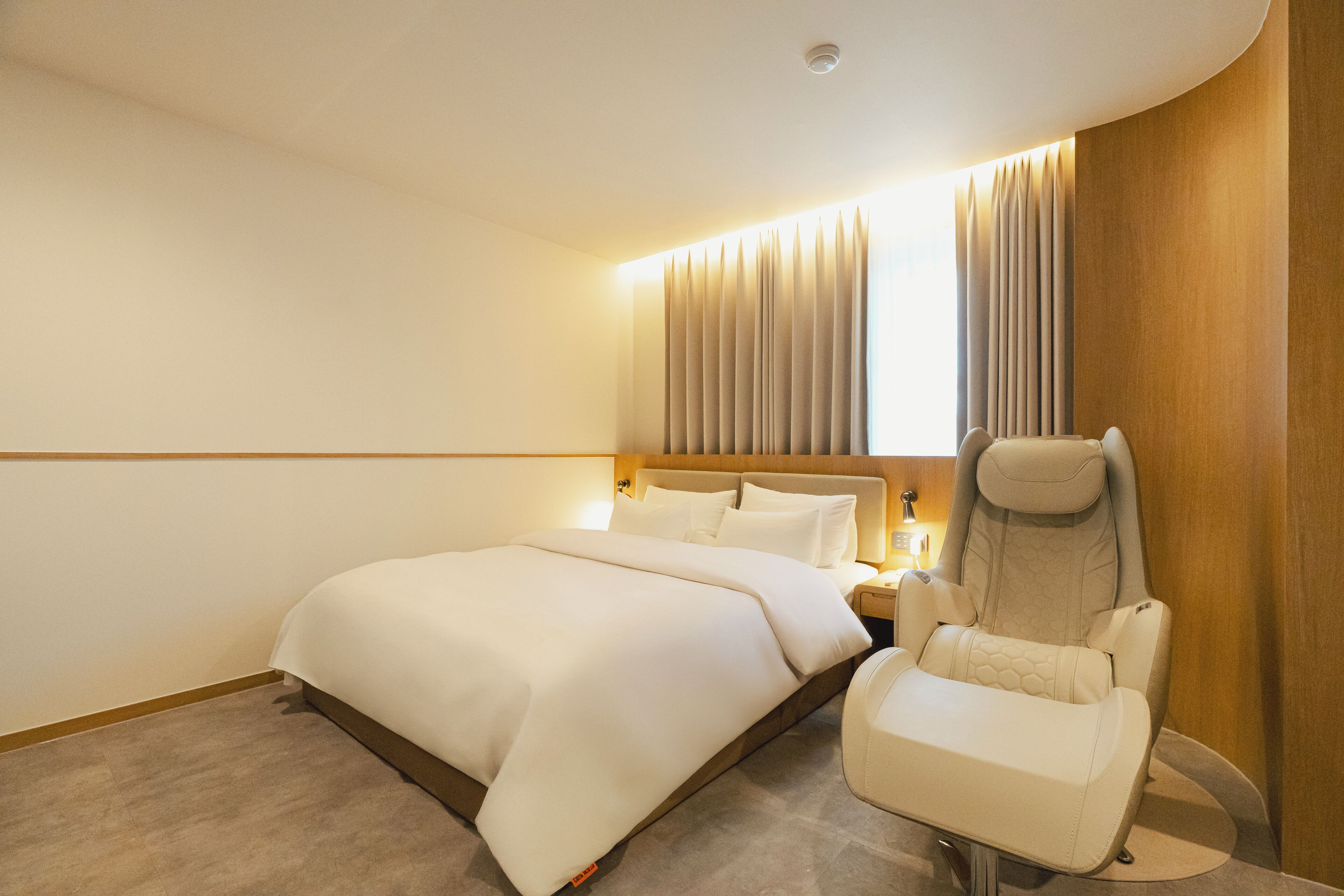 Quarto de hotel com uma cama grande, uma mesa de cabeceira, uma cadeira e uma janela com cortinas.