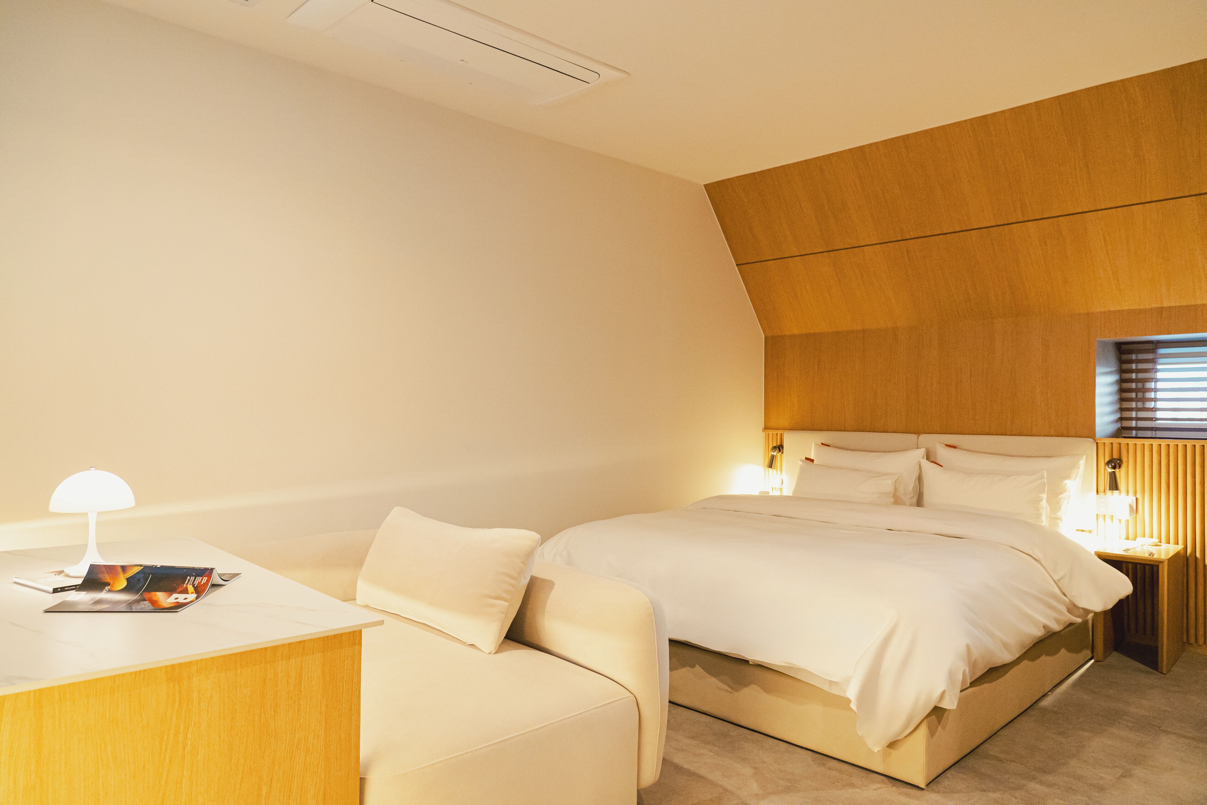 Um quarto moderno com uma cama grande, uma mesa de cabeceira com abajur, e uma pequena escrivaninha com abajur e revistas.