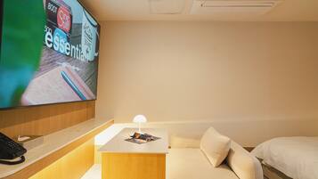 Quarto de hotel com televisão na parede, escrivaninha de madeira com luminária, mesinha e sofá com almofada.