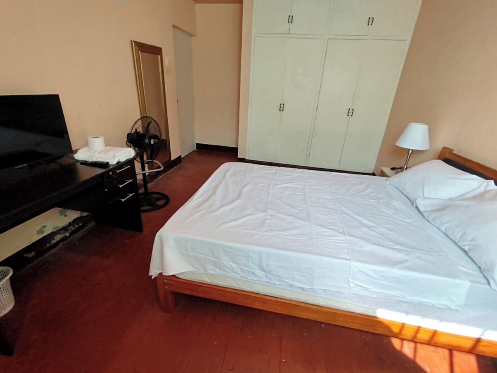 Quarto de hotel com cama, televisão, guarda-roupa e secretária com abajur.