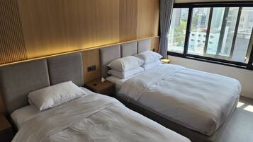 Quarto de hotel com duas camas, cabeceira de madeira, mesa de cabeceira com abajur e janela com vista para a cidade.