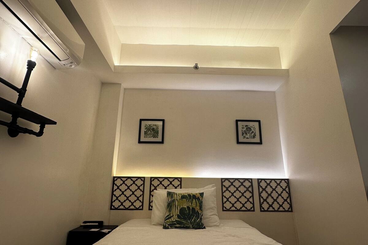 Quarto de hotel com uma cama, uma mesinha de cabeceira, uma prateleira na parede e duas fotografias emolduradas.