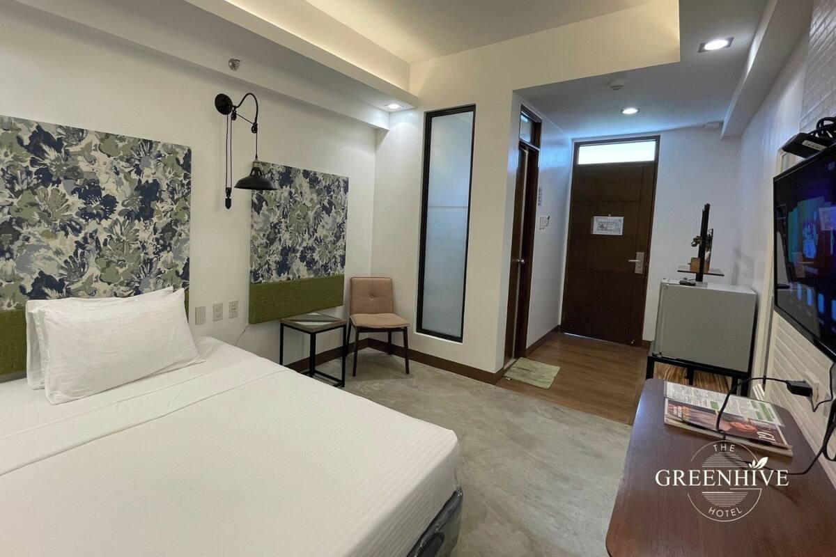 Quarto de hotel com cama, poltrona, escrivaninha, televisão e porta.