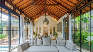 Ampla sala de estar com um sofá modular grande, mesa de centro de madeira e janelas amplas com vista para piscina e vegetação.
