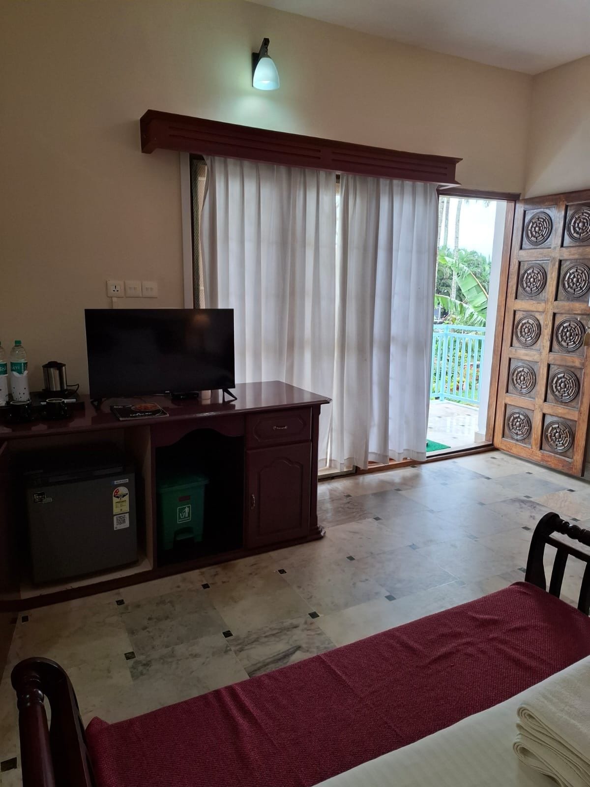 Quarto com cama, escrivaninha com televisão, pequeno frigorífico, janela com cortinas e porta com painel decorativo.