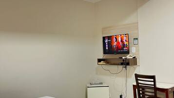 Quarto de hotel com cama, escrivaninha, cadeira, televisão presa à parede e um pequeno frigorífico.