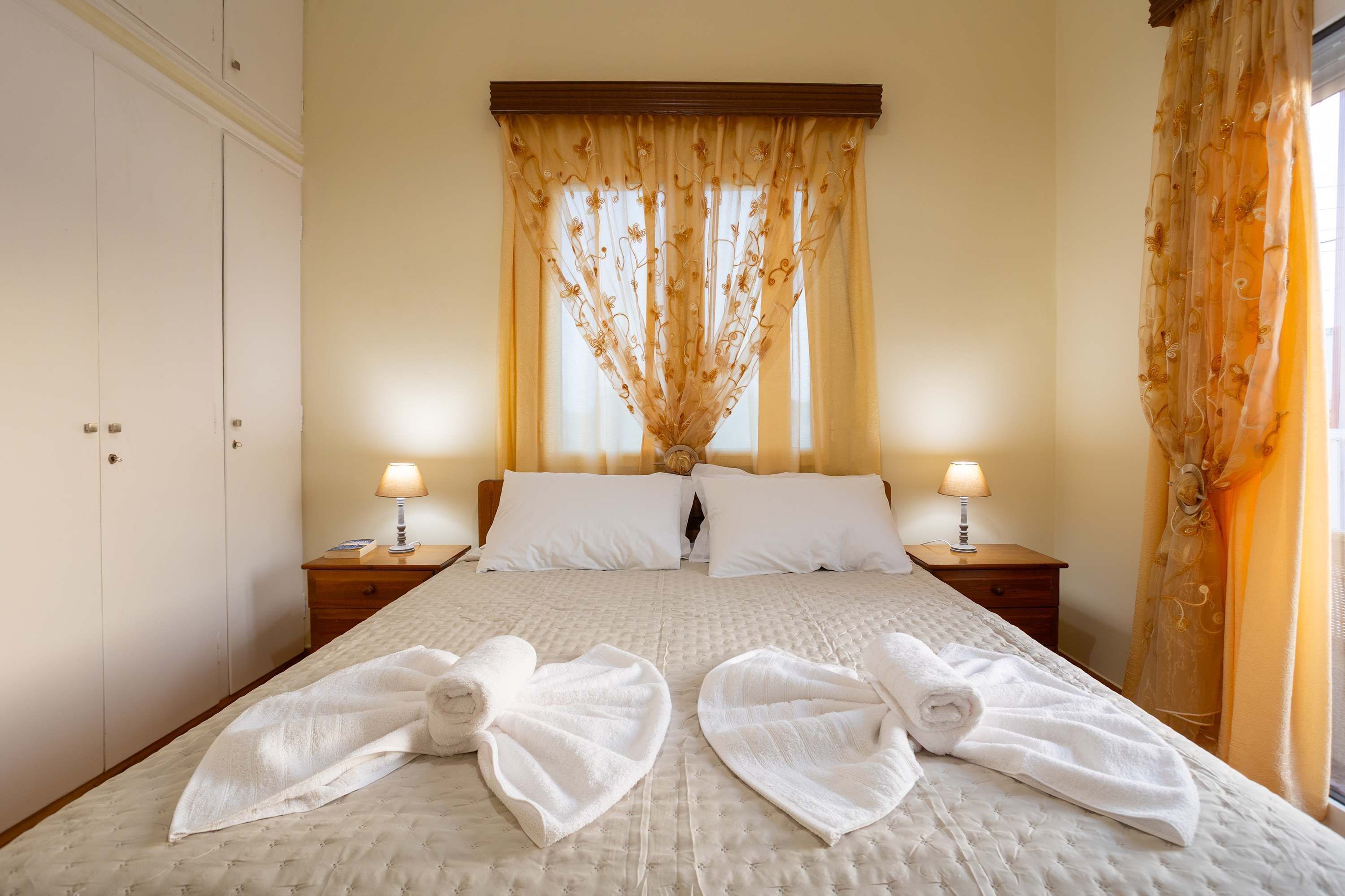 Cama bem arrumada com duas almofadas, mesa de cabeceira com abajur e janela com cortinas finas.