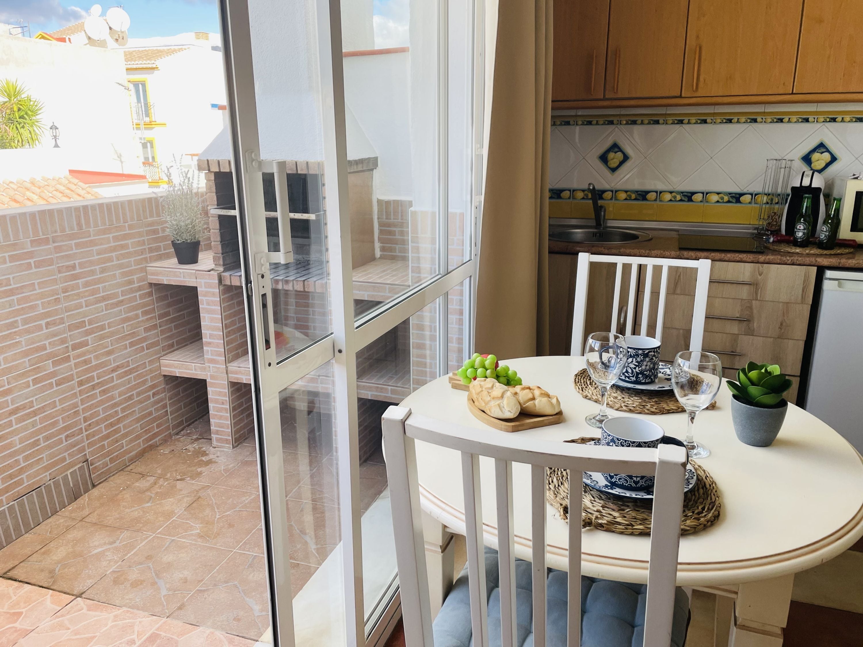 Uma pequena área de refeições com mesa branca, uma cadeira, um vaso de planta e vista para o exterior através de uma porta de vidro.
