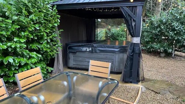 Uma mesa com tampo de vidro, quatro cadeiras de madeira, uma banheira de hidromassagem e um gazebo num jardim.