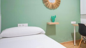 Quarto de hotel com cama, espelho redondo, pequeno cacto decorativo e escrivaninha com cadeira.
