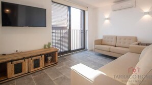 Uma sala de estar moderna com uma televisão de tela plana, um sofá bege, um móvel de entretenimento de madeira e uma porta de vidro deslizante que dá para uma varanda.