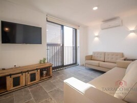 Uma sala de estar moderna com uma televisão de tela plana, um sofá bege, um móvel de entretenimento de madeira e uma porta de vidro deslizante que dá para uma varanda.