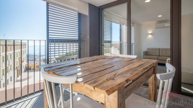 Uma varanda com mesa e cadeiras de madeira, janelas amplas e vista para edifícios.