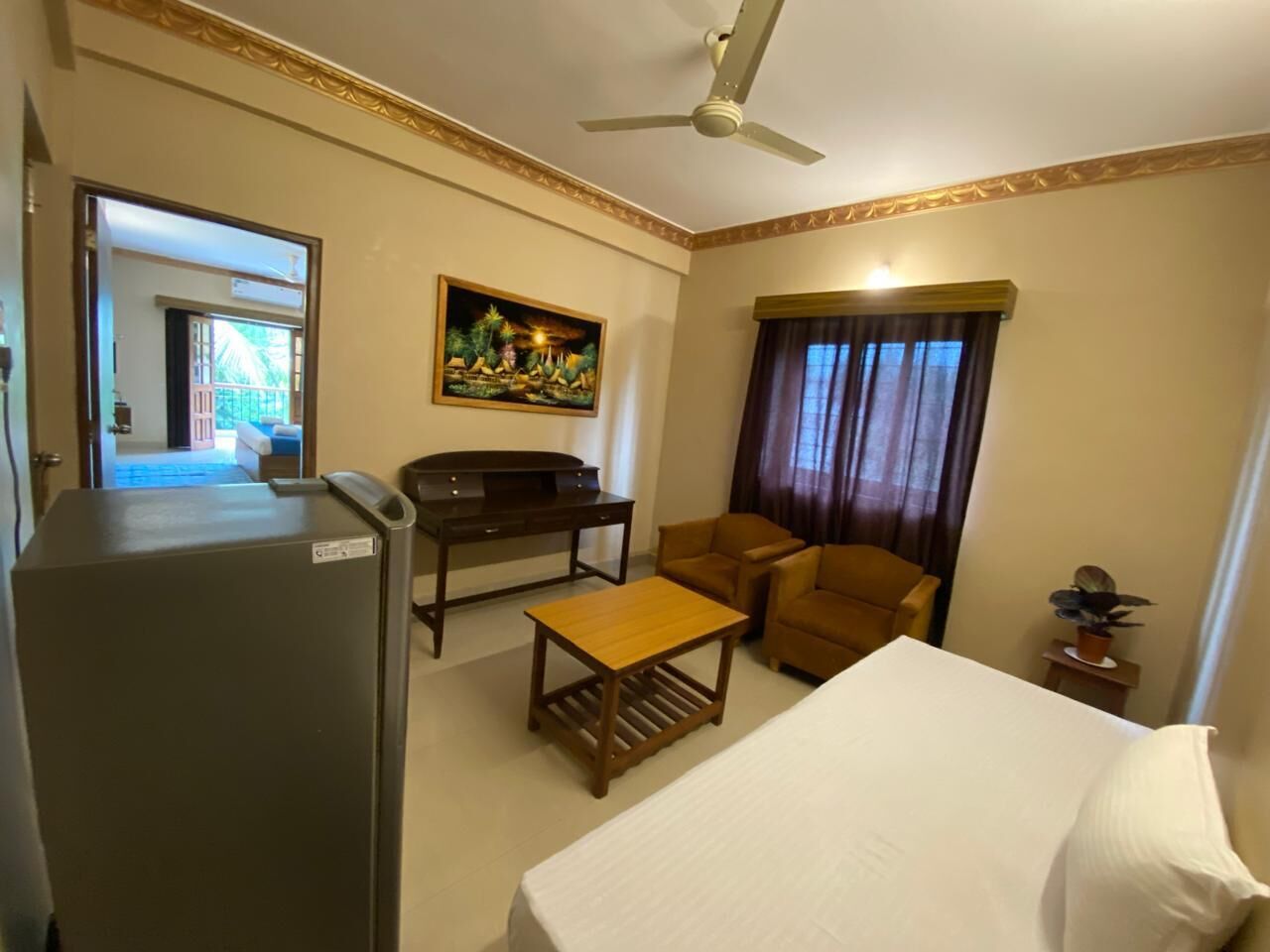 Quarto de hotel com cama, escrivaninha, cadeira, mesa pequena, frigorífico, ventoinha no teto, um quadro e janela com cortinas.