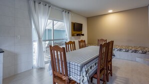 Uma sala de jantar com mesa posta para seis, cadeiras de madeira, televisão fixada na parede e uma janela com cortinas.