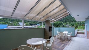 Um alpendre coberto com mesa e cadeiras, churrasqueira e vista para uma paisagem verde.