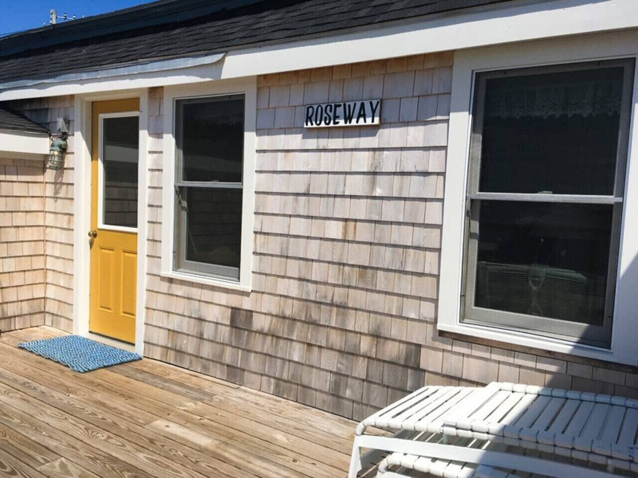 Uma casa com um letreiro 'ROSEWAY', um deck de madeira e uma porta.