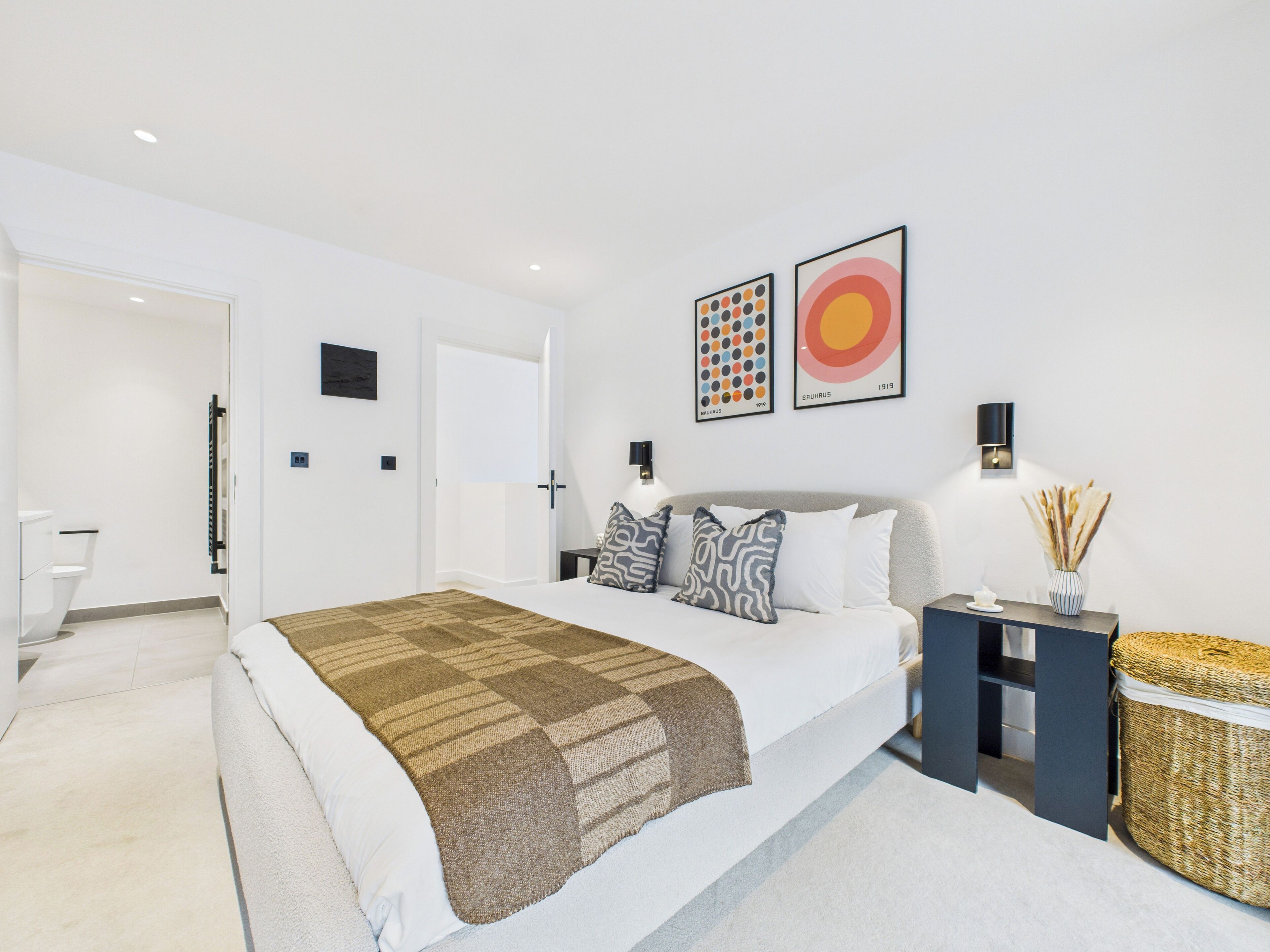 Quarto moderno com cama grande, mesa de cabeceira, luminária na parede e duas pinturas abstratas.