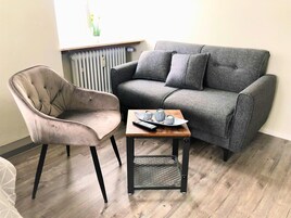Uma sala de estar com um sofá cinzento, uma poltrona estofada e uma pequena mesa de centro de madeira, com um controle remoto e objetos decorativos.