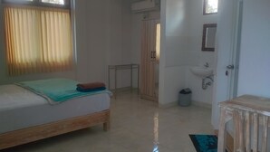 Quarto com cama, mesa de cabeceira, janela com cortinas, porta, mesinha e lixeira.
