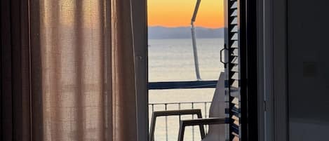 Quarto com cama, uma cadeira e vista para o mar ao pôr do sol.
