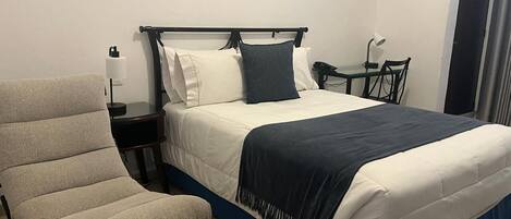 Quarto de hotel com cama, cadeira, mesa de cabeceira e escrivaninha com abajur.