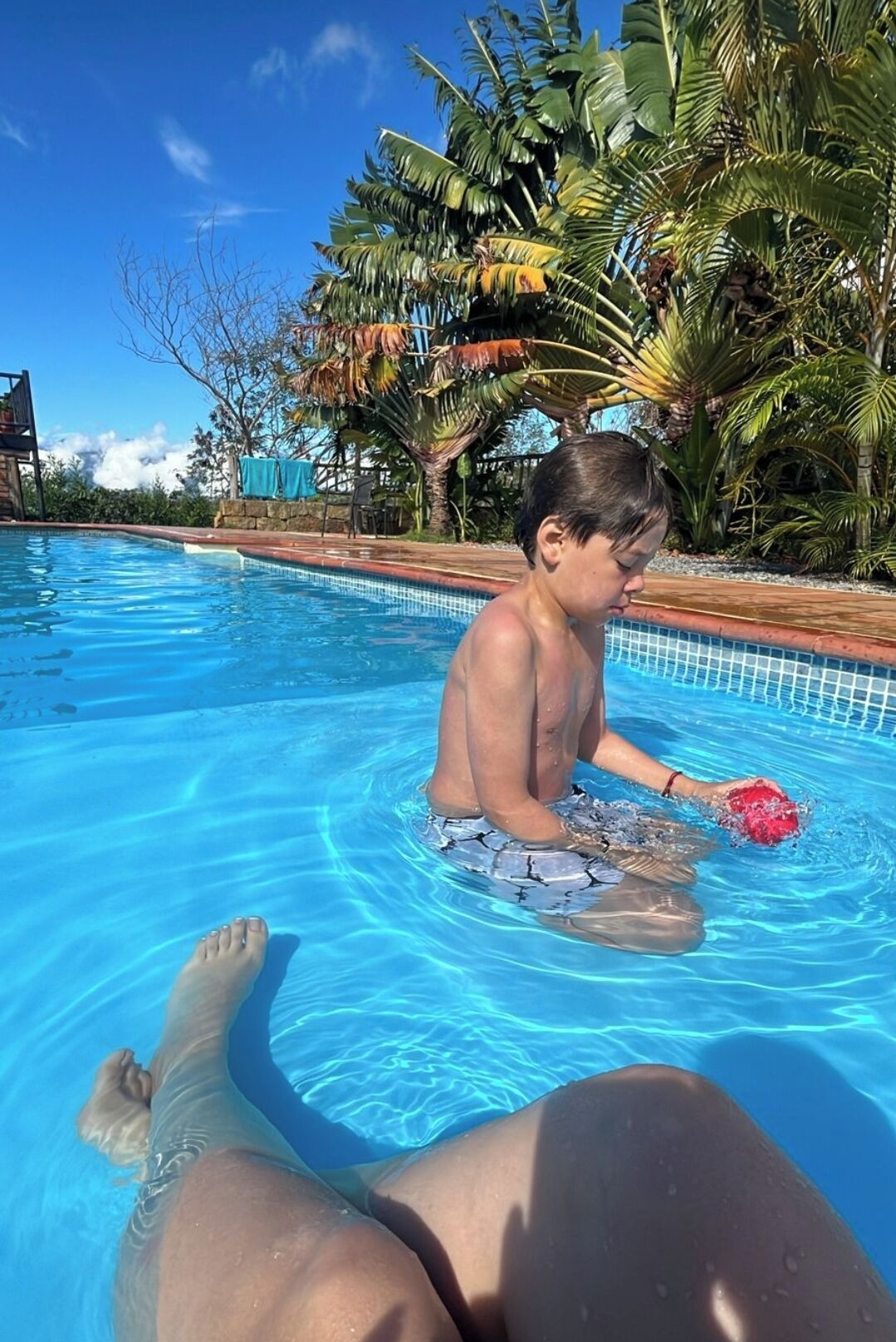 Uma pessoa em uma piscina com água azul cristalina, rodeada por plantas tropicais e um céu limpo.