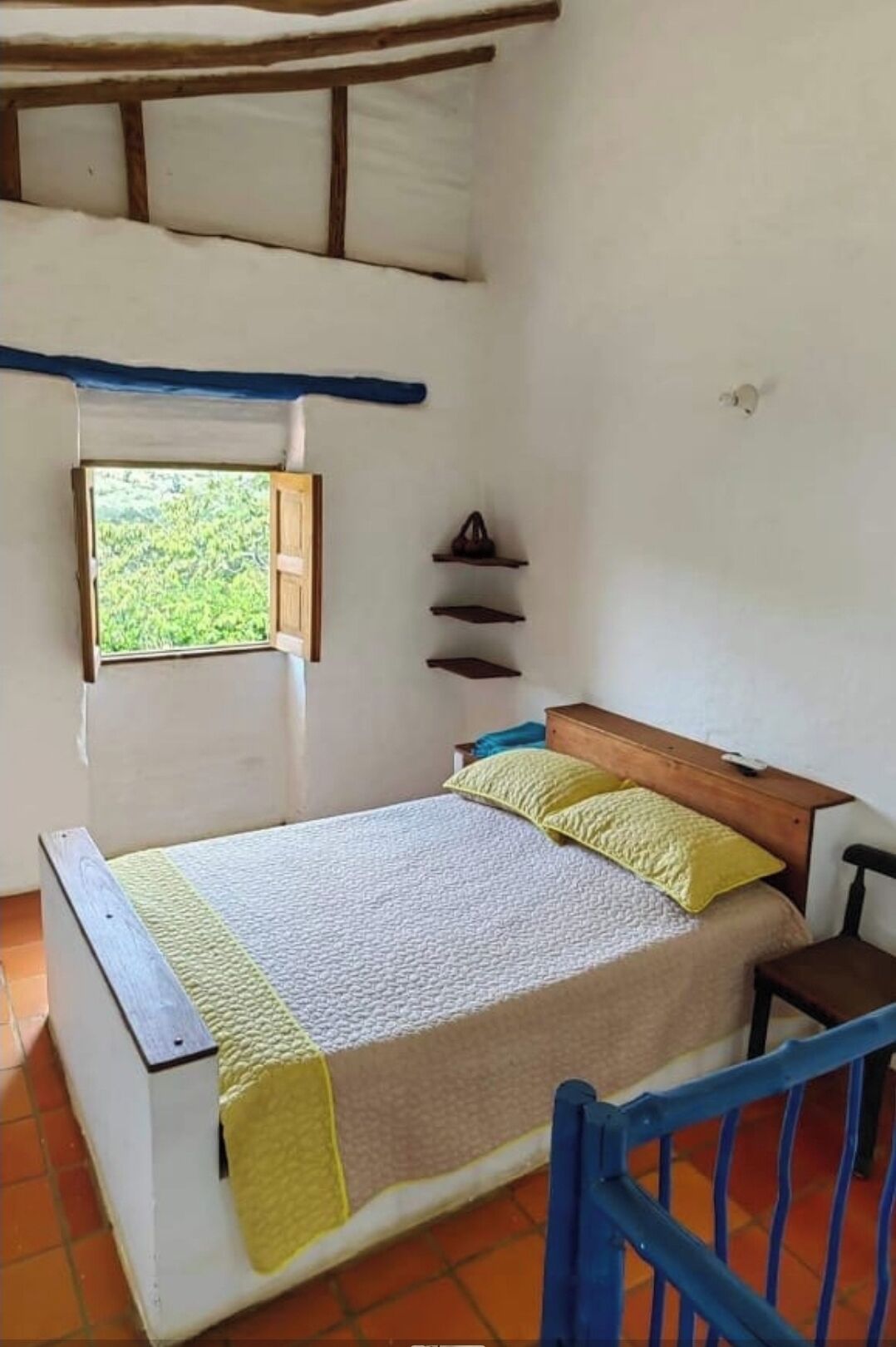 Quarto individual com uma cama, janela, cabeceira de madeira e uma pequena estante.