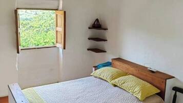 Quarto individual com uma cama, janela, cabeceira de madeira e uma pequena estante.