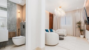 Uma sala de estar moderna com um sofá branco, uma mesa de centro redonda e um candelabro. Há um banheiro com vaso sanitário branco e um armário de madeira.