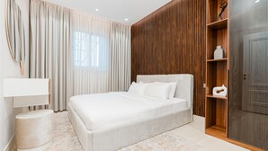Quarto moderno com uma cama grande, luminária de cabeceira, guarda-roupa de madeira e janela com cortinas leves.